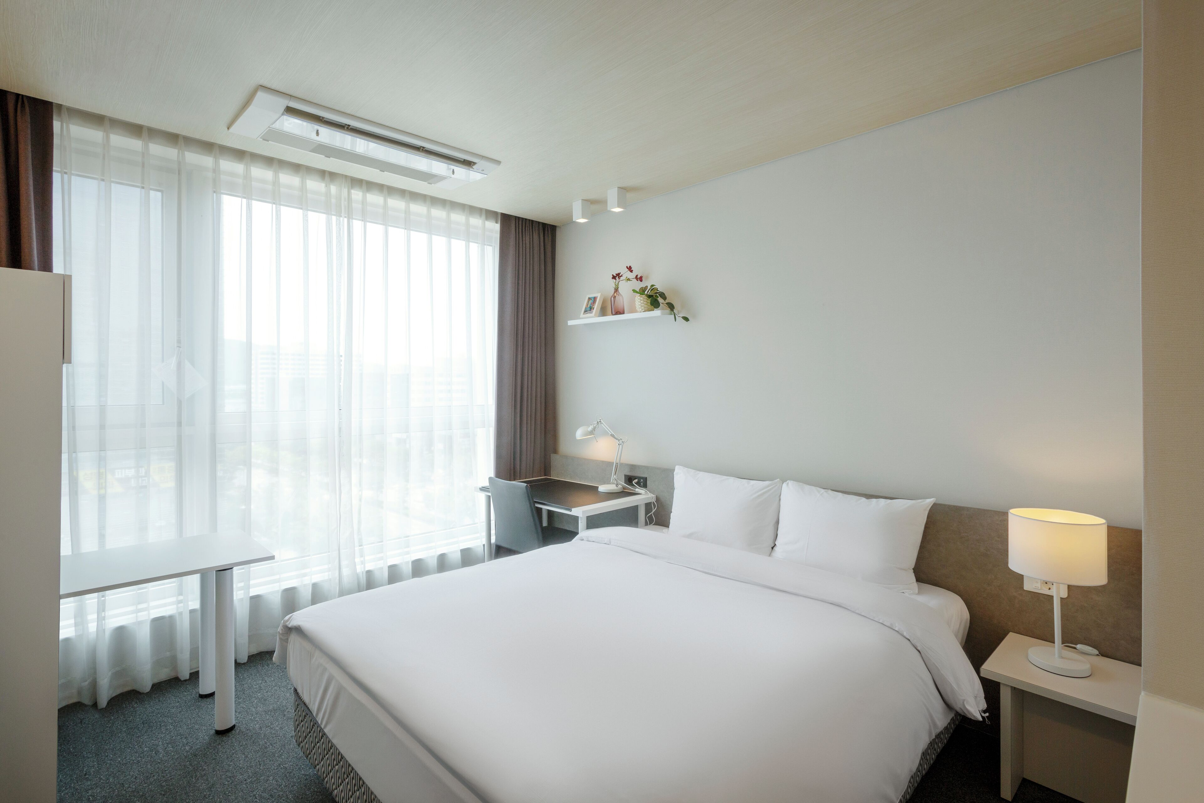 Quarto de hotel com uma cama grande, uma escrivaninha, uma cadeira, uma mesa de cabeceira e uma prateleira fixada na parede com um vaso e uma foto.