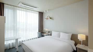 Quarto de hotel com uma cama grande, uma escrivaninha, uma cadeira, uma mesa de cabeceira e uma prateleira fixada na parede com um vaso e uma foto.