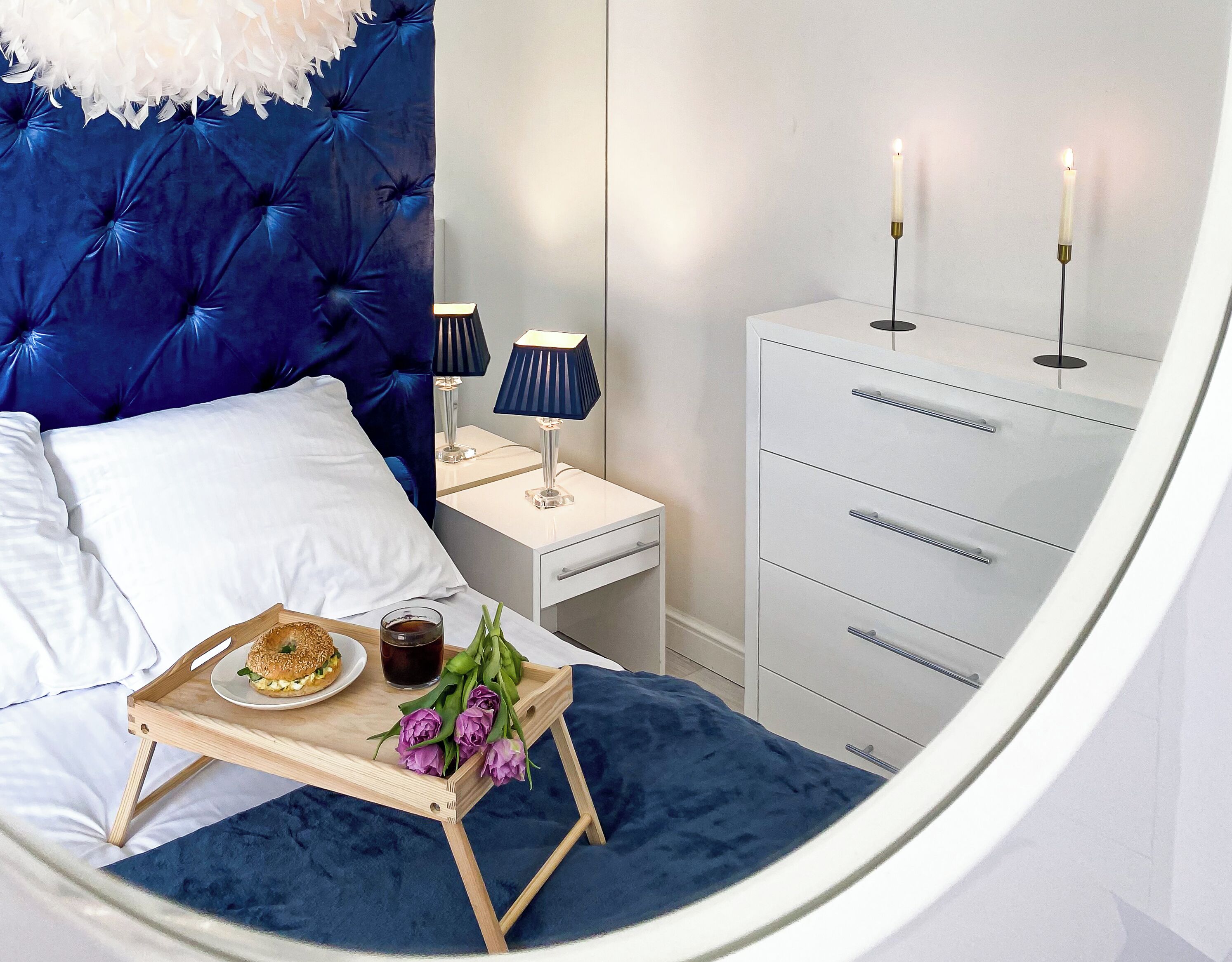 Quarto com cabeceira estofada, mesa de cabeceira com luminárias, cômoda e bandeja com comida e flores.