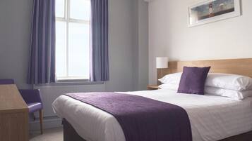 Quarto de hotel com uma cama, uma cadeira, uma escrivaninha e uma janela com cortinas.