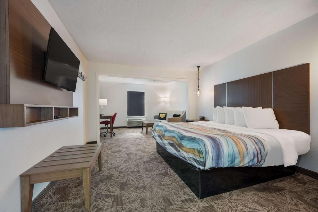 Quarto de hotel com cama, escrivaninha, cadeira, televisão e janela.