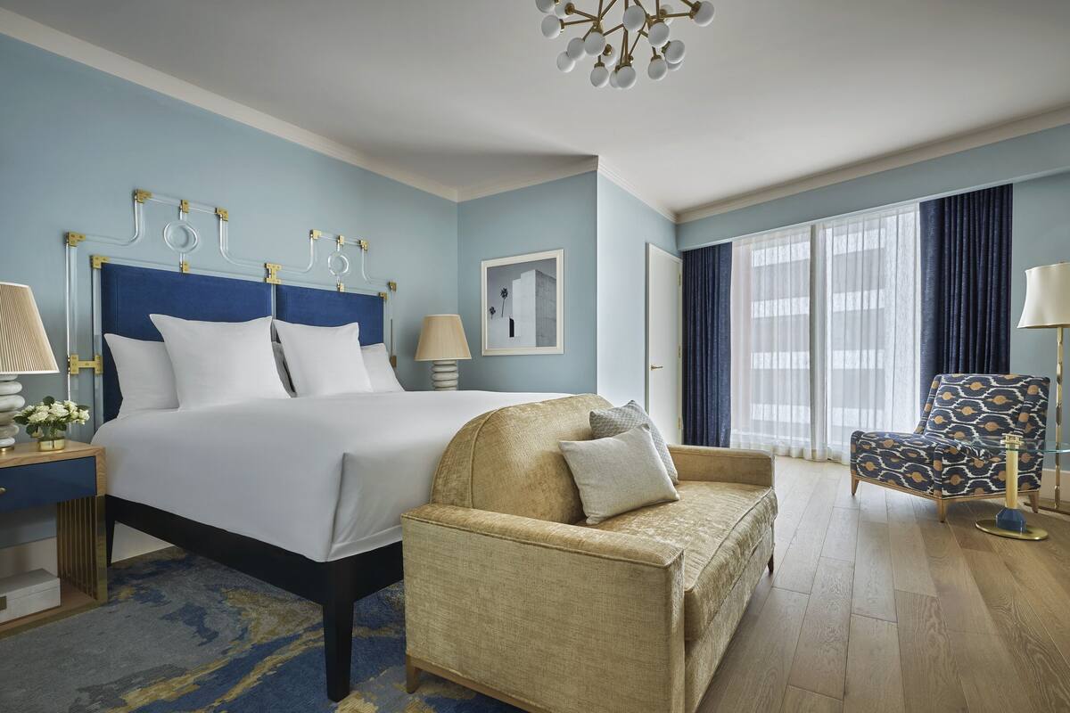 Quarto de hotel com uma cama, um sofá, uma cadeira, uma mesa de cabeceira e uma luminária.