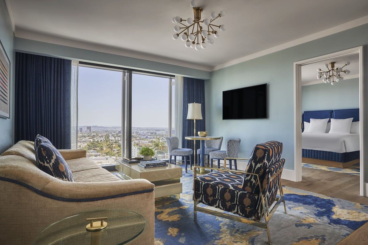 Quarto de hotel com uma janela grande, um sofá, uma poltrona estampada, uma televisão e uma cama.