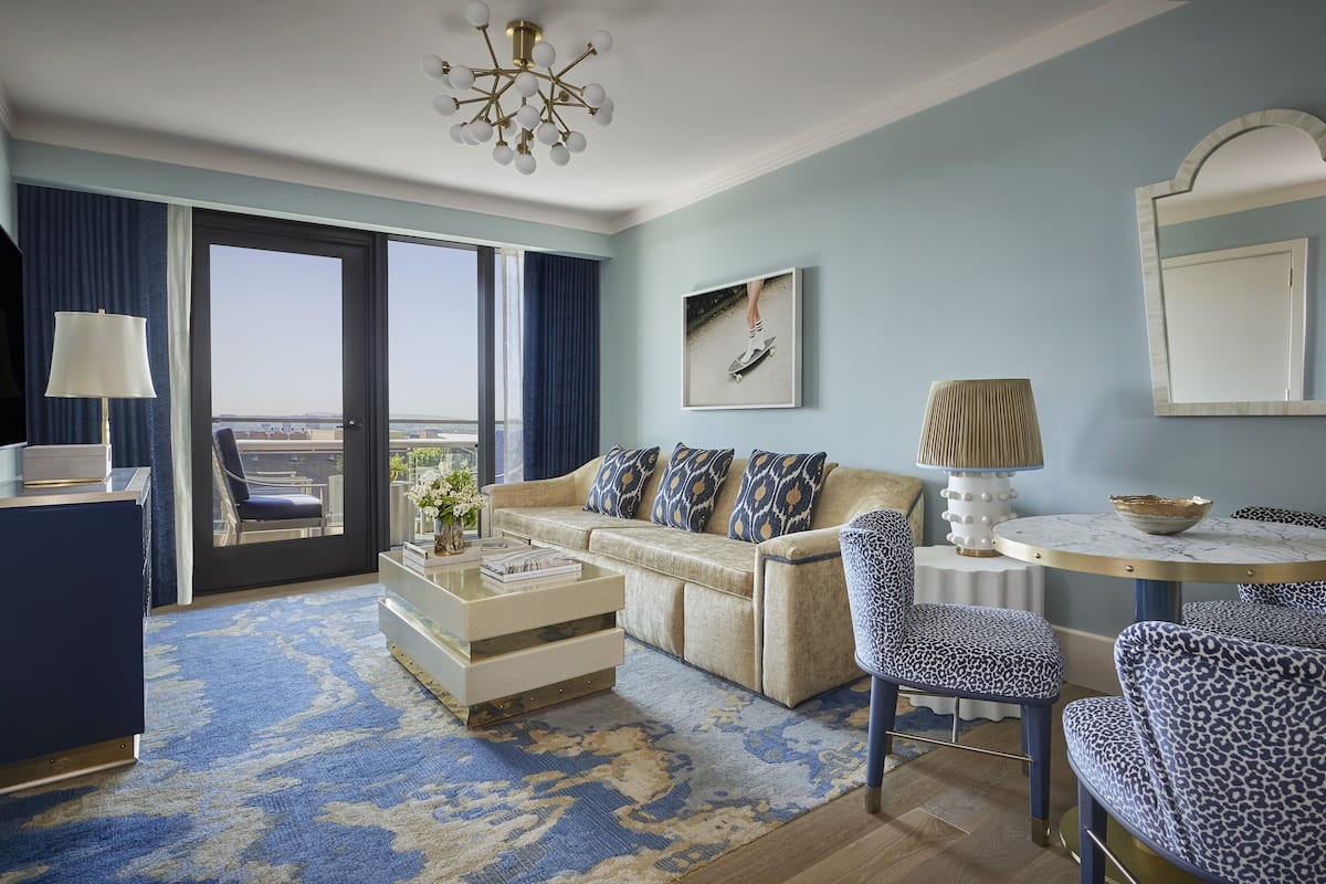 Quarto de hotel moderno com sofá, poltronas, mesa de centro e mesa de jantar. Há uma janela grande com vista para a cidade.