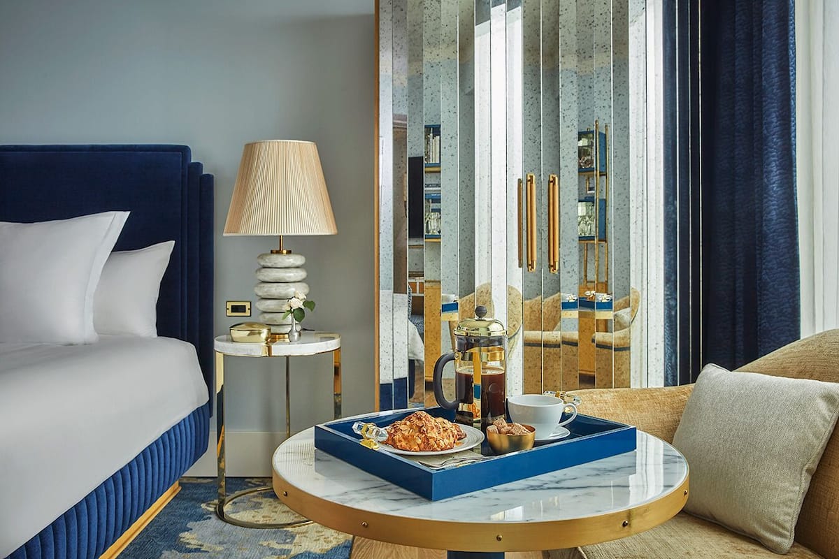 Quarto de hotel com uma cama, uma mesa de centro redonda com itens de café da manhã, um abajur e um espelho refletindo o quarto.