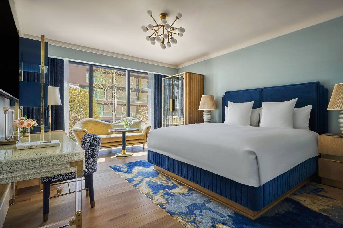 Quarto de hotel com uma cama grande, uma escrivaninha com abajur, uma cadeira e vista para o exterior.
