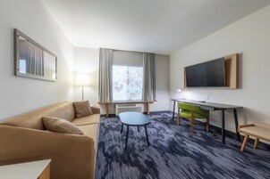 Quarto de hotel com sofá, mesa pequena, escrivaninha com cadeira, televisão de tela plana, janela com cortinas e radiador.