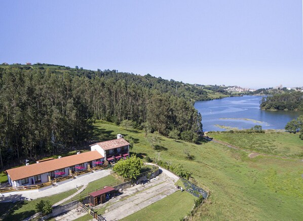 Um hotel com um edifício de telhado vermelho, área de pátio com guarda-sóis e vista para um lago e colinas arborizadas.