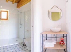 Casa de banho com chuveiro em cabine de vidro, sanita, lavatório com espelho e prateleira com toalhas e uma caixa.