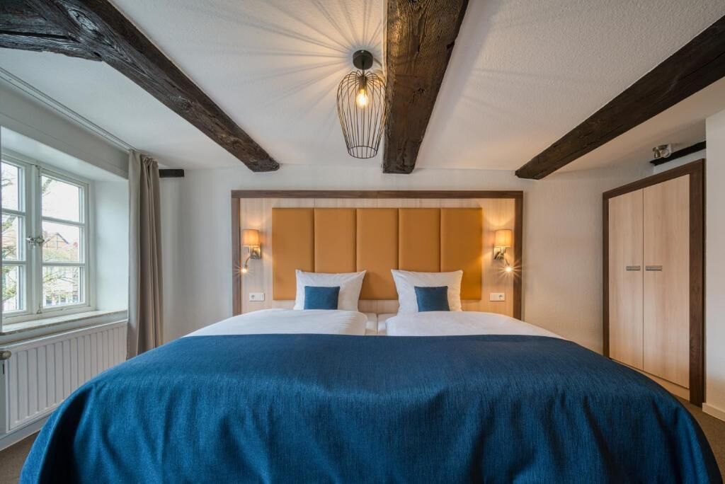 Uma cama grande com colcha azul, cabeceira de madeira e duas luminárias laterais.