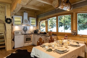 Cozinha acolhedora de uma cabana de madeira com mesa de jantar posta para refeição, grandes janelas e decoração rústica.
