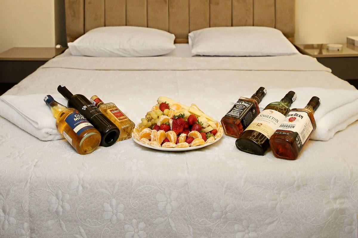 Cama bem arrumada com almofadas, uma travessa de frutas e garrafas de bebidas alcoólicas.