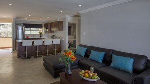 Uma sala de estar moderna com um sofá de couro preto, uma mesa de centro com uma fruteira e um vaso com flores. Há uma área de cozinha com geladeira, banquetas de bar e uma mesa de jantar.