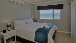 Quarto com cama, mesa de cabeceira, luminária, janela com vista para o mar e ar condicionado.