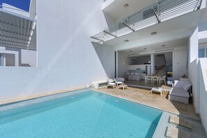 Uma área moderna à beira da piscina, com piscina transparente, mobiliário de exterior branco e uma área de refeições envidraçada.