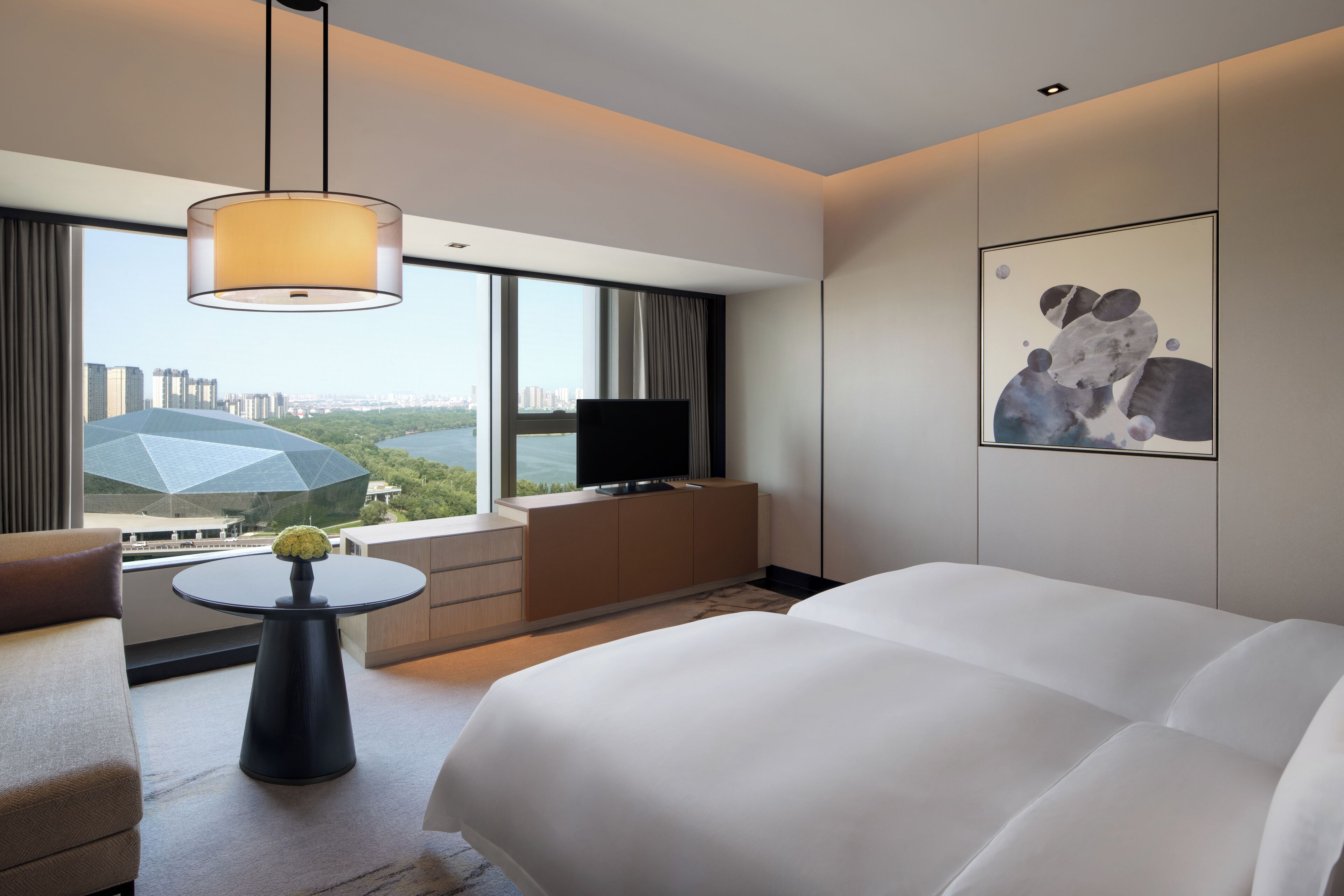 Quarto de hotel moderno com uma cama grande, um sofá, uma pequena mesa com um objeto decorativo, uma televisão em um móvel e uma janela ampla com vista para a cidade.