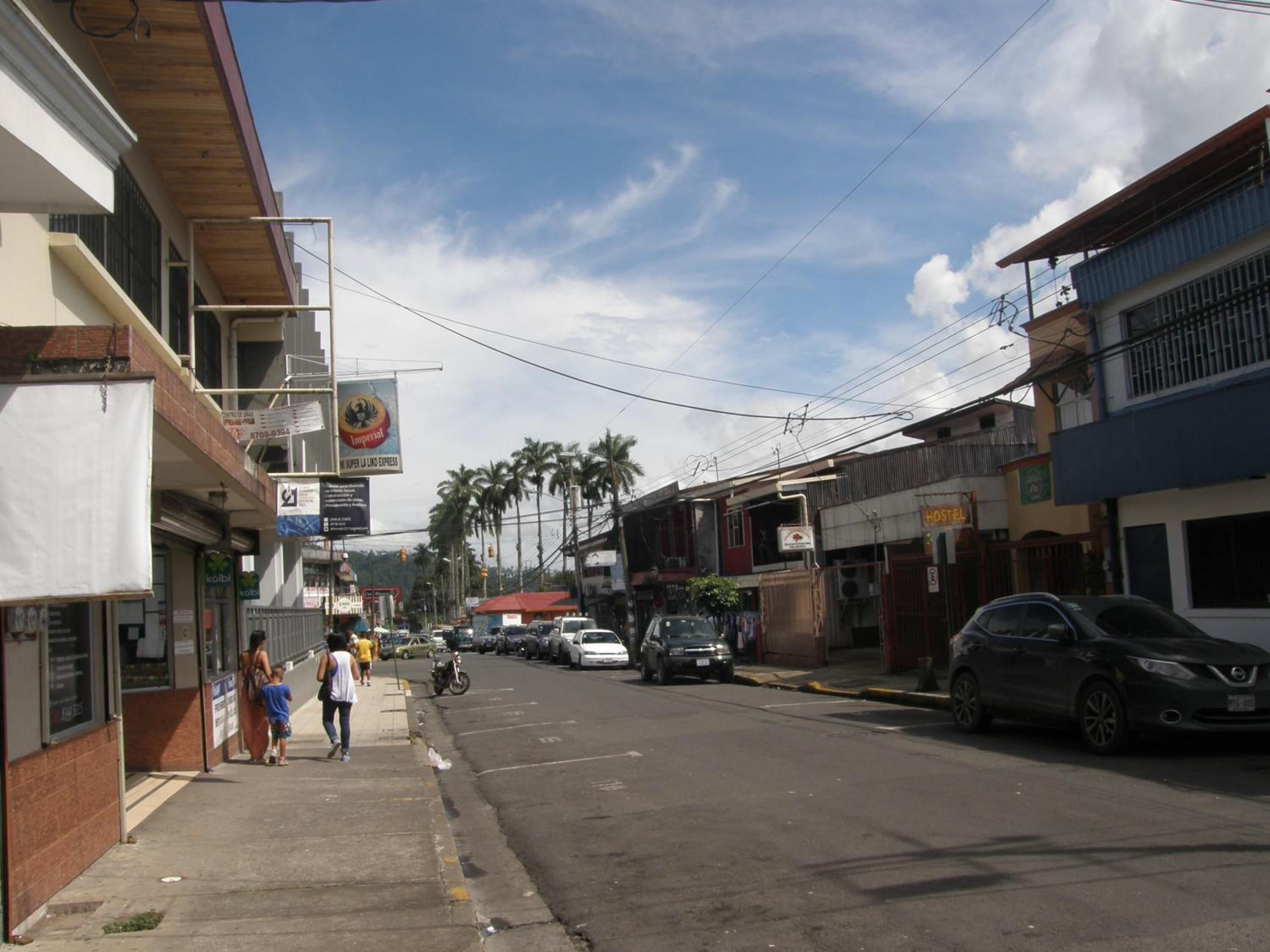 Vista da rua com edifícios, carros estacionados e pedestres.