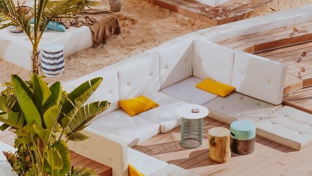 Uma área à beira da piscina com assentos acolchoados brancos, um deck de madeira e uma praia de areia.