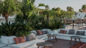 Um terraço com área de lounge, sofás brancos, mesas de madeira redondas e diversas plantas.