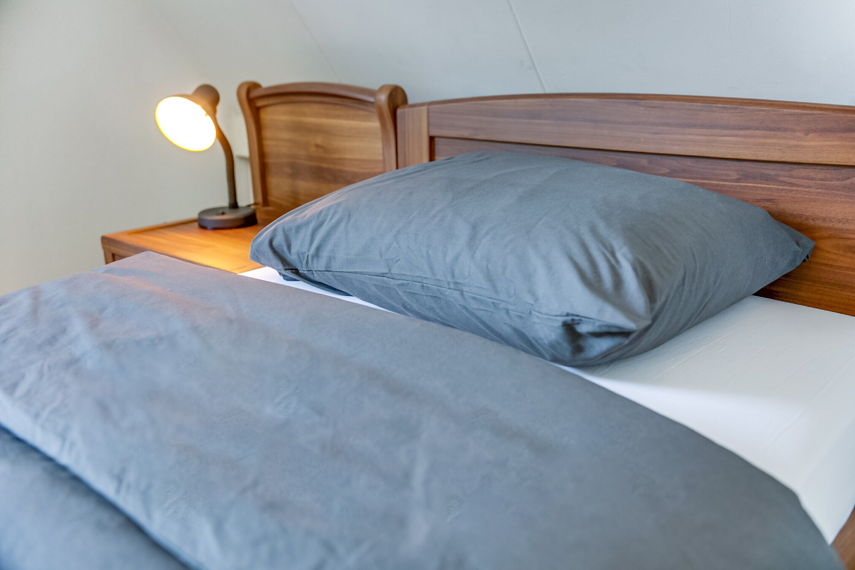 Cama bem arrumada com travesseiro cinza e lençol branco, cabeceira de madeira e mesa de cabeceira com abajur.