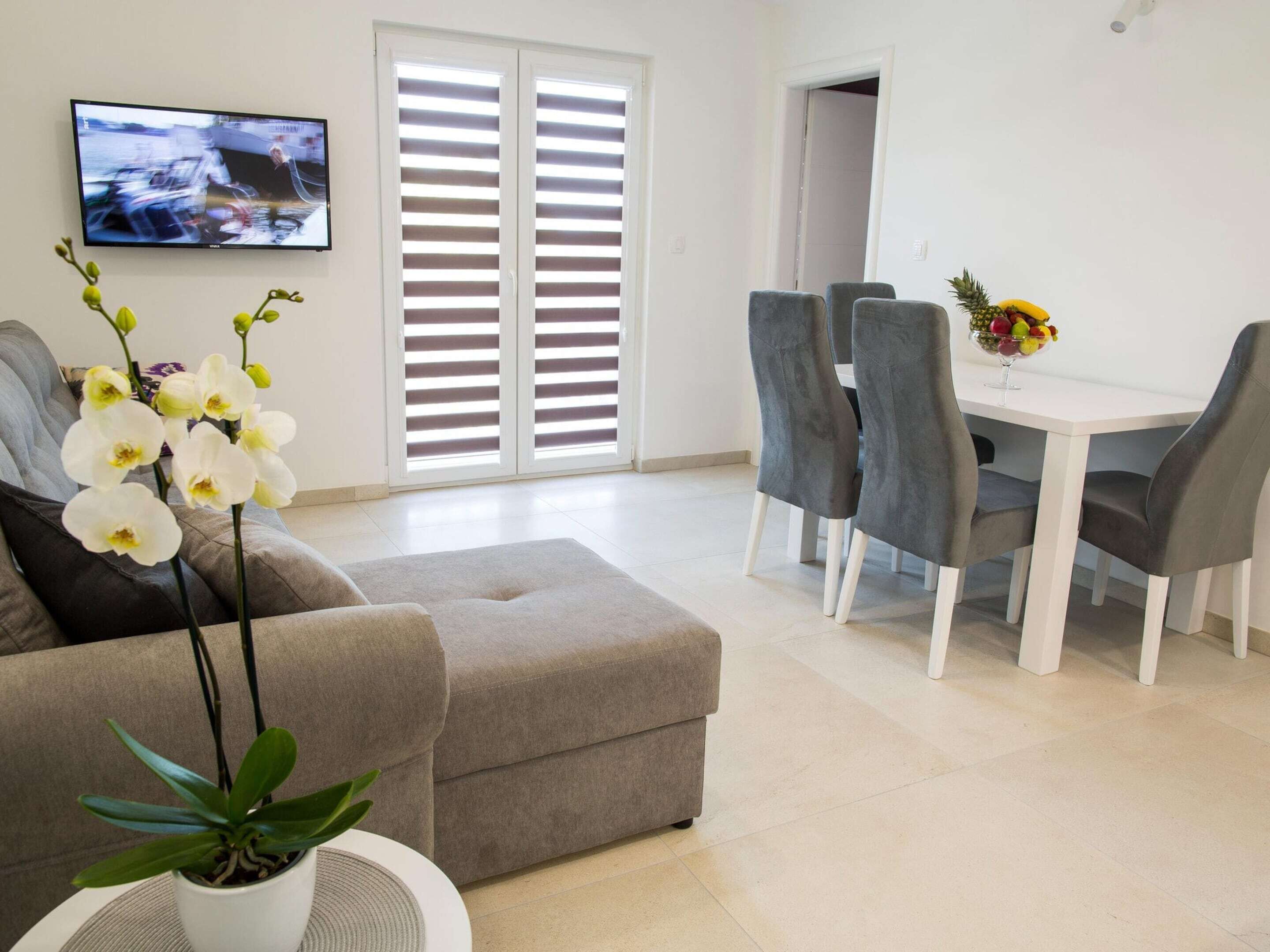 Uma sala de estar moderna com um sofá cinzento, uma mesa de jantar com quatro cadeiras e uma TV fixada na parede.