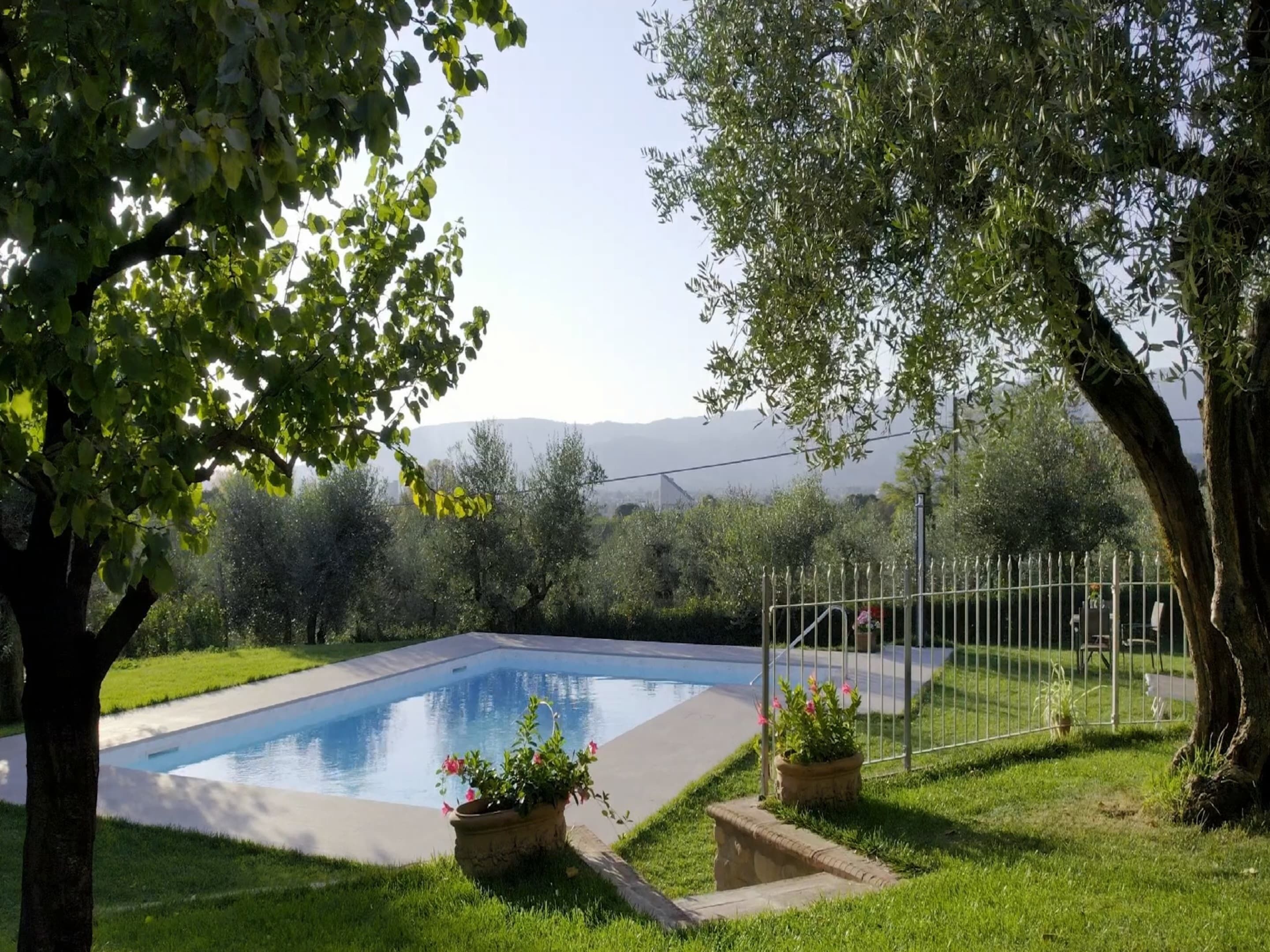 Uma piscina cercada por grama e árvores, com cerca de metal e vista para montanhas ao longe.
