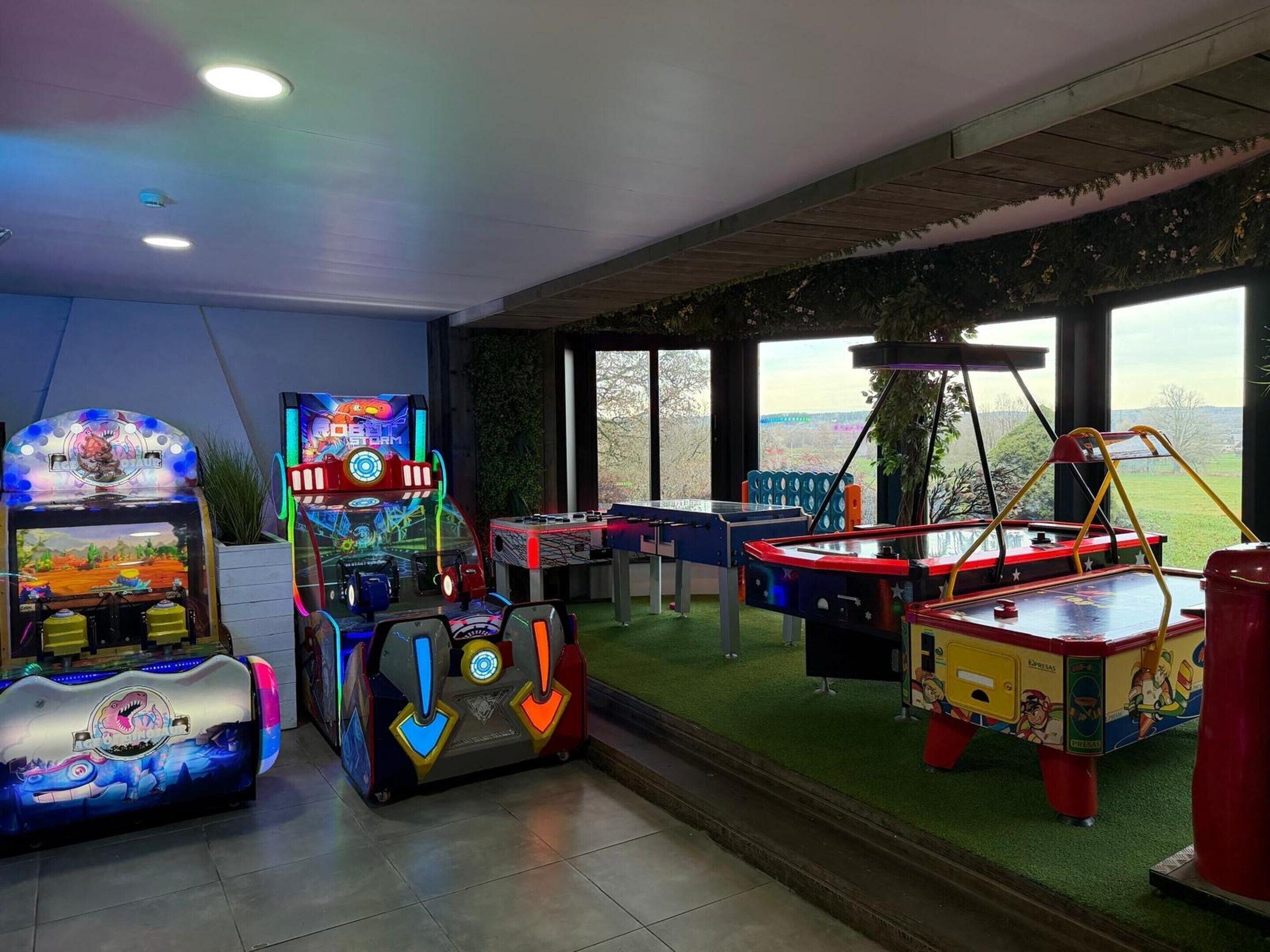 Uma área de lazer interna com jogos de fliperama, mesas de pebolim e vista para o exterior através de grandes janelas.
