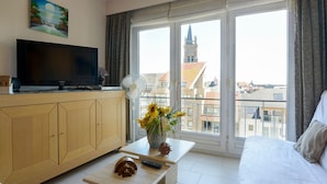 Quarto de hotel com uma janela grande, uma televisão sobre um móvel de madeira, uma mesa pequena com um vaso de flores e uma cama.