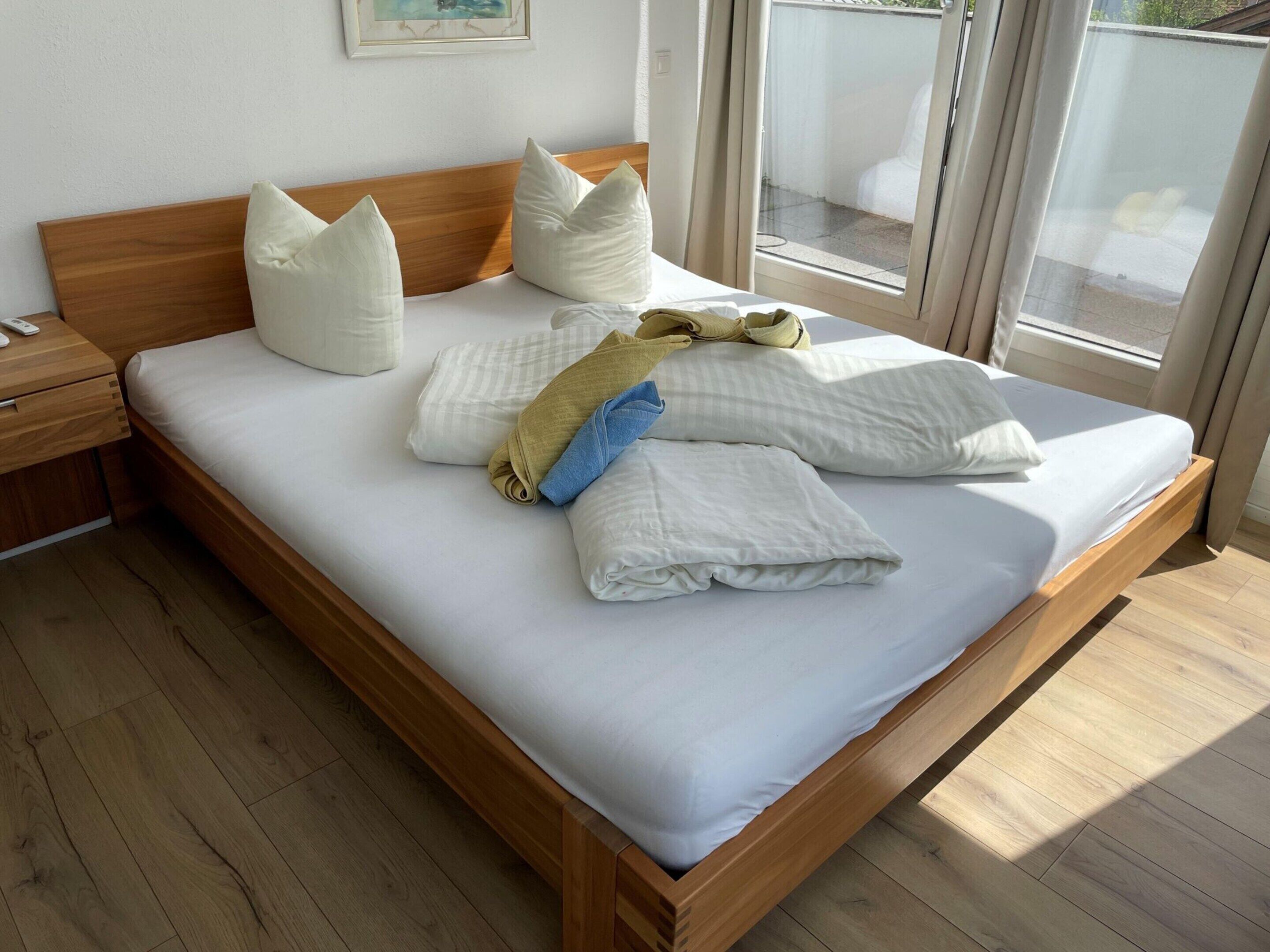 Cama bem arrumada com lençóis brancos e várias almofadas, cabeceira de madeira, mesa de cabeceira de madeira com controle remoto e uma janela com cortinas.