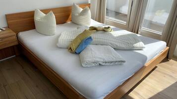 Cama bem arrumada com lençóis brancos e várias almofadas, cabeceira de madeira, mesa de cabeceira de madeira com controle remoto e uma janela com cortinas.