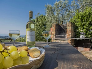 Uma garrafa de vinho Monteolivo, dois copos de vinho branco, uma cesta de uvas e uma mesa rústica ao ar livre com um forno de tijolos ao fundo.
