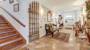 Um corredor espaçoso com escada de madeira, um portão decorativo e uma coleção de cadeiras e armas antigas.