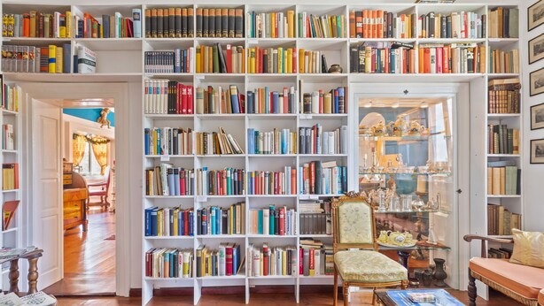 Uma biblioteca ampla com várias estantes cheias de livros, uma poltrona confortável e uma mesa de centro com livros.