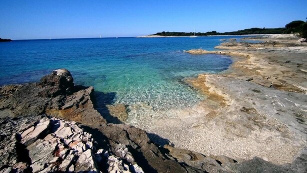 Uma costa rochosa com águas azul-claras e um veleiro ao longe.