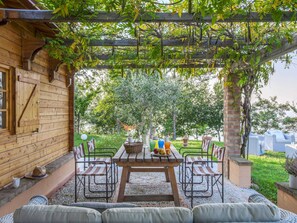 Uma cabana de madeira com alpendre coberto, com uma mesa e cadeiras, cercada por vegetação e uma rede.