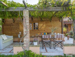 Uma cabana rústica de madeira com um espaço de varanda com mesa e cadeiras, um sofá de vime e uma planta pendente.