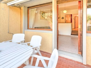 Um pátio com uma mesa e cadeiras brancas, uma janela com cortinas e vista para o interior do edifício.