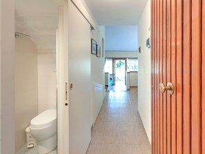 Um corredor com um banheiro, uma porta de madeira e piso de azulejos.
