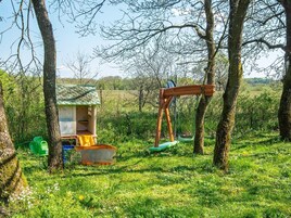 Uma casinha de madeira rústica com telhado verde, balanço e um pequeno galpão em área gramada cercada por árvores.