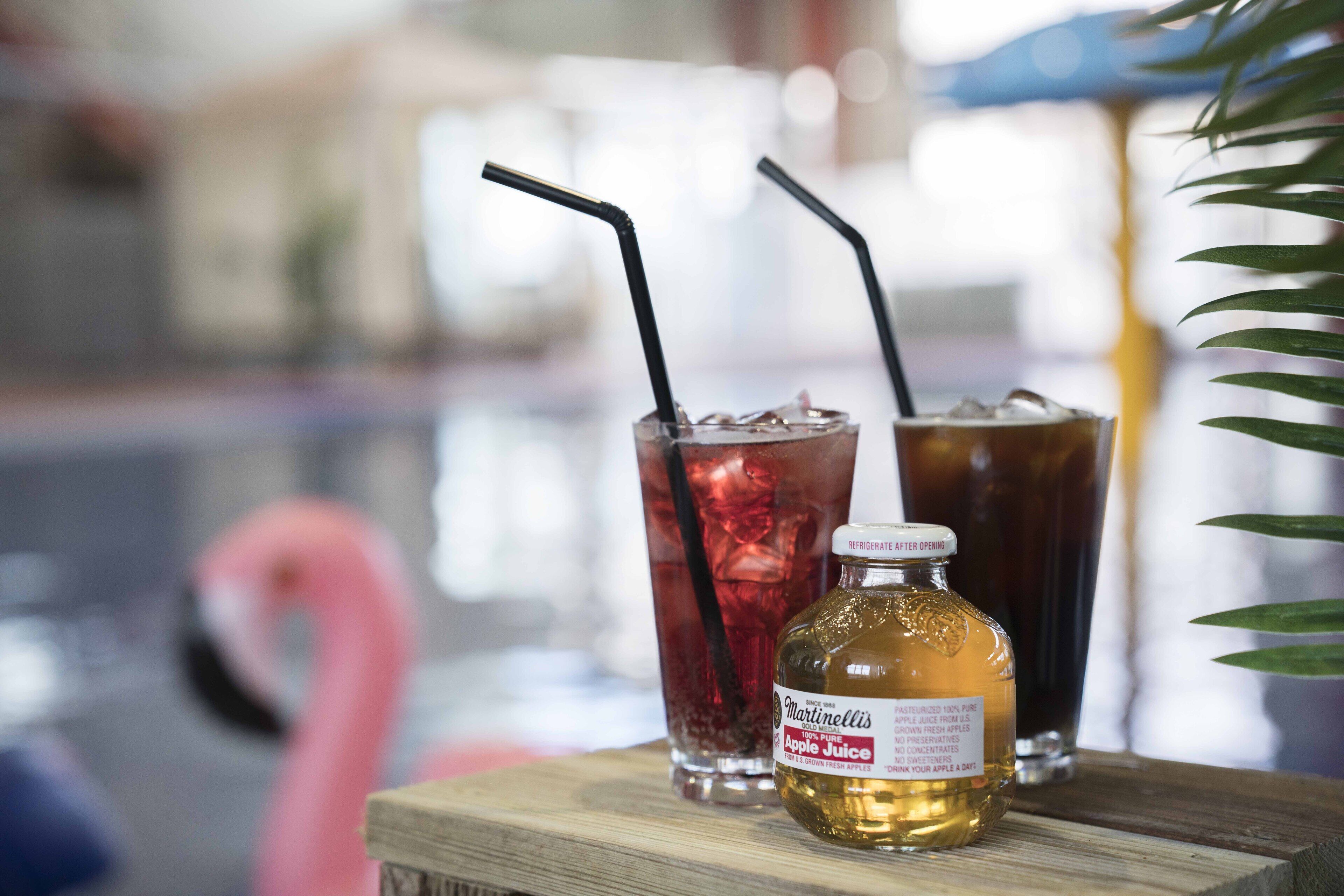 Duas bebidas geladas com canudos pretos, uma garrafa de suco de maçã e uma decoração de flamingos cor-de-rosa.