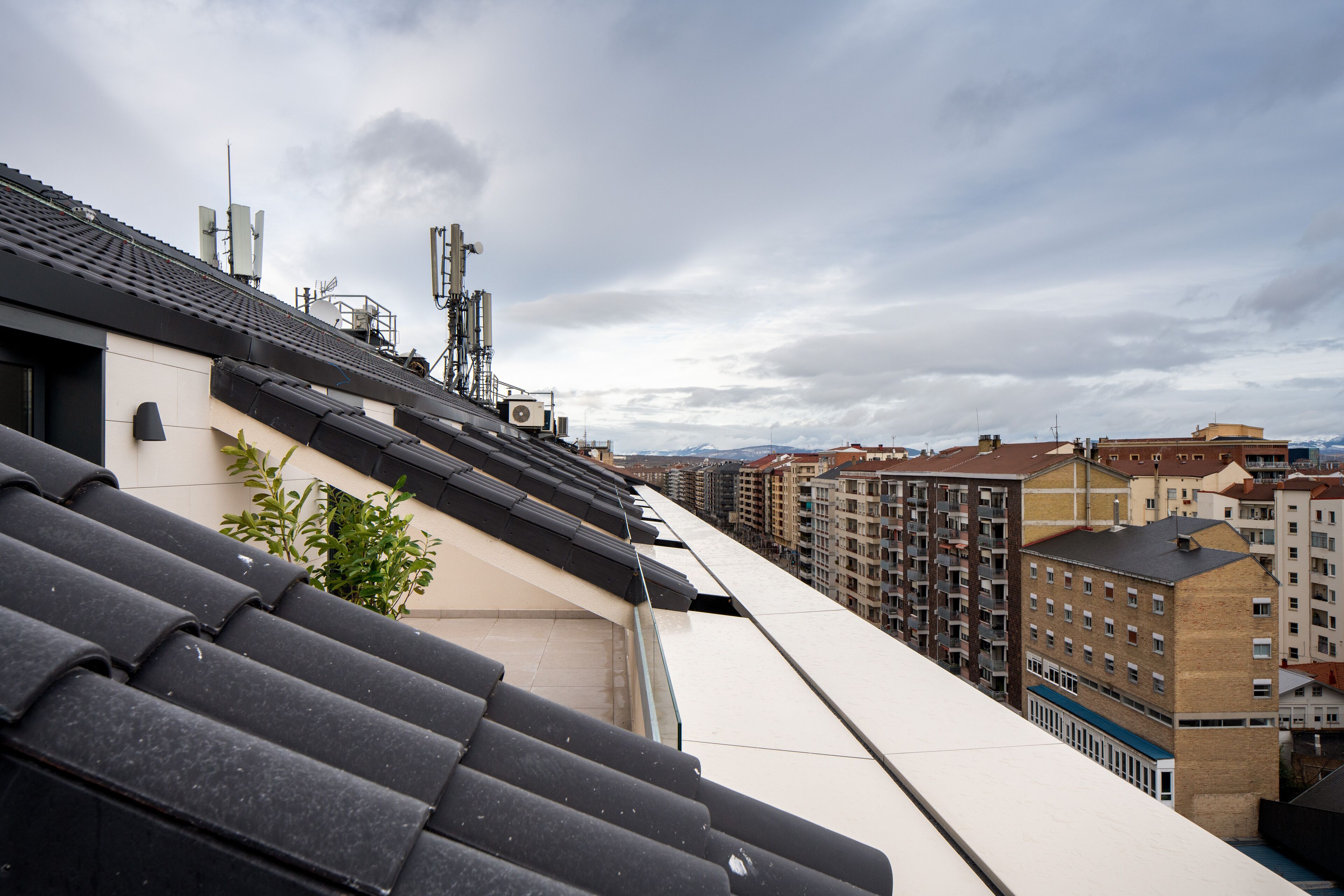 Terraço com telhado inclinado, antenas parabólicas e vista dos edifícios ao redor.