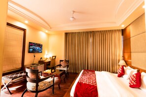 Quarto de hotel com cama, escrivaninha, cadeiras, televisão e uma grande janela com cortinas.