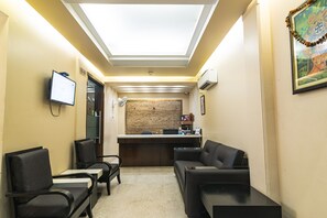 Uma área de recepção com balcão, cadeiras, televisão de tela plana, luminária no teto, ar-condicionado fixado na parede e um quadro emoldurado.
