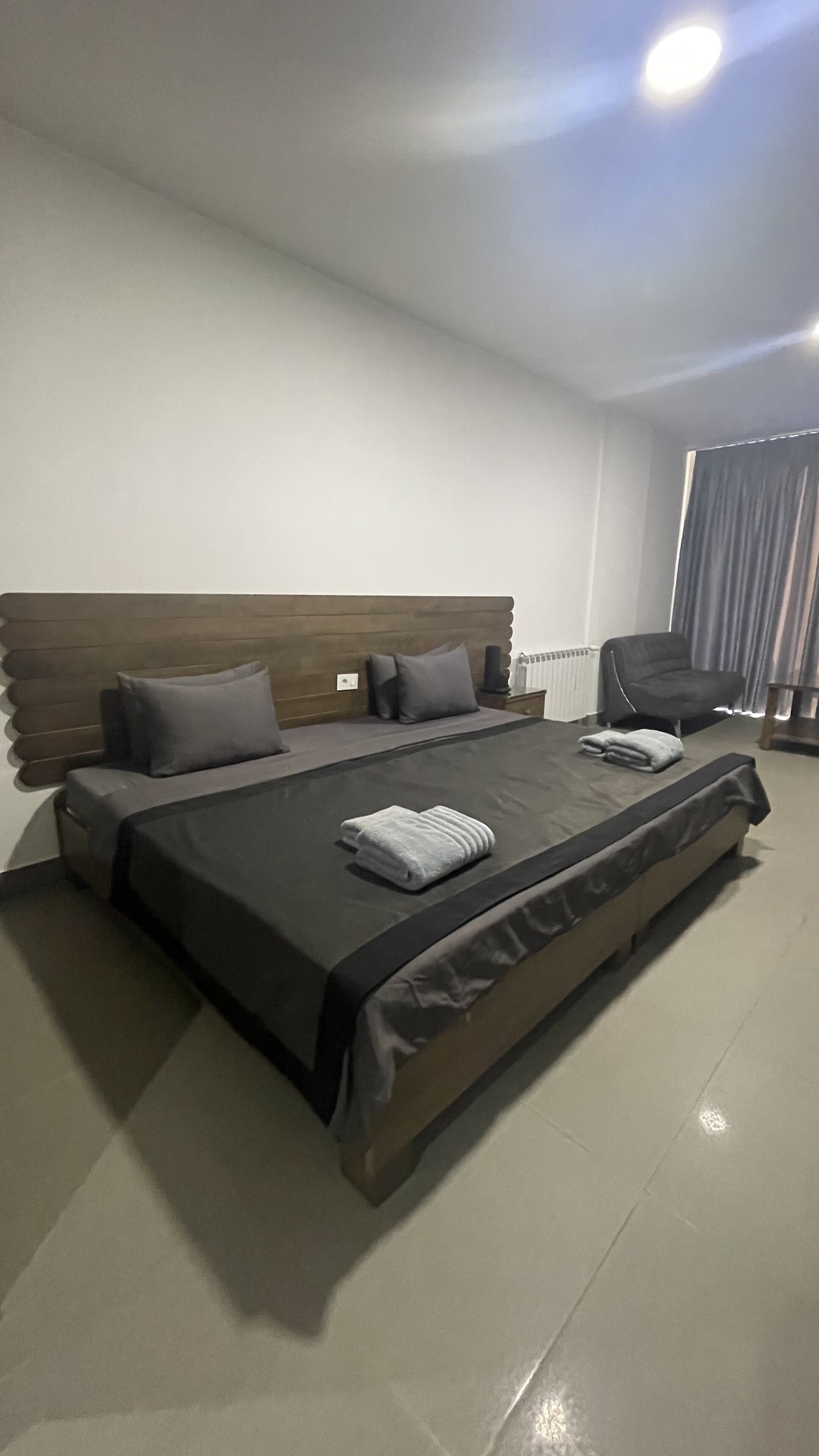 Quarto de hotel moderno com uma cama grande, cabeceira de madeira, uma cadeira e vista para o exterior.