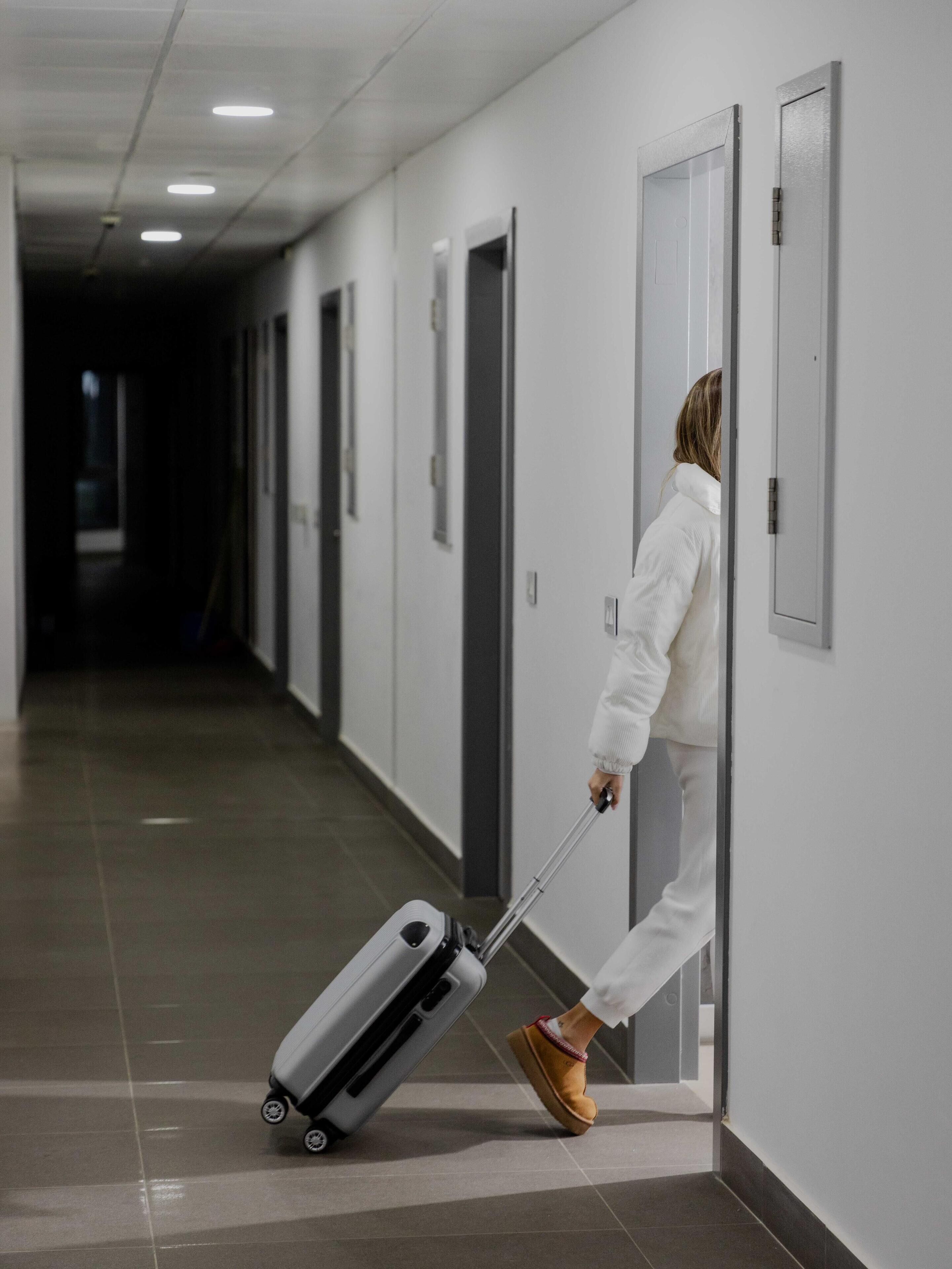 Uma pessoa caminhando por um corredor com uma mala, portas à direita e piso de azulejos.