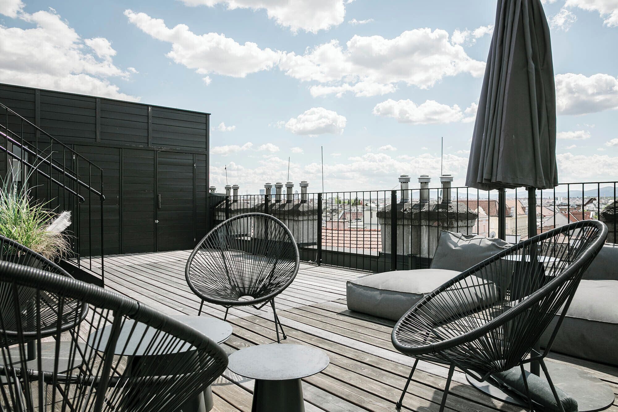 Um terraço no telhado com guarda-corpo de metal preto, cadeiras de vime, uma mesa redonda e um guarda-sol cinza.