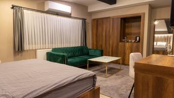 Quarto de hotel com uma cama, um sofá verde, uma mesa pequena branca, um guarda-roupa de madeira e uma escrivaninha com luminária.
