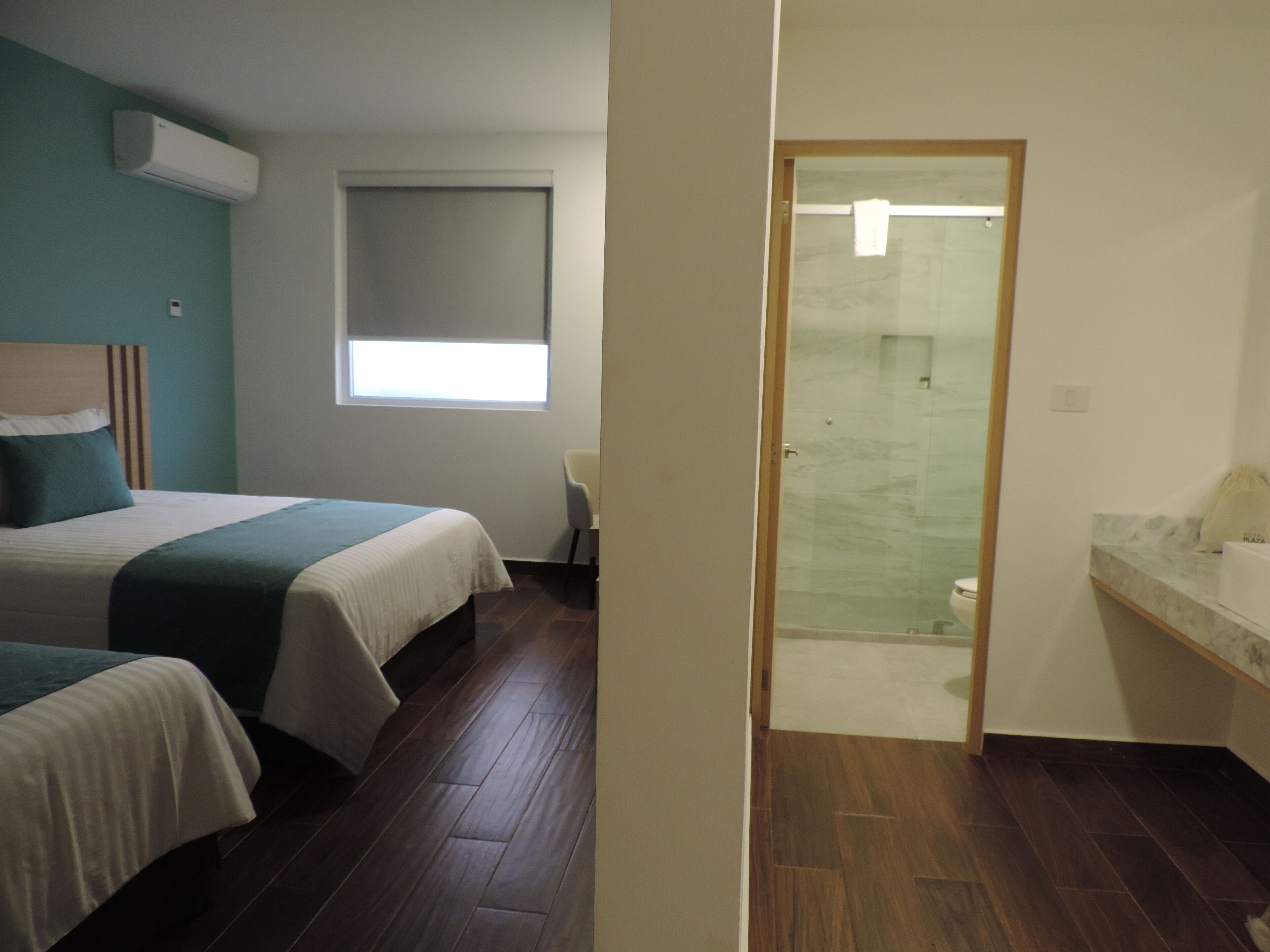 Quarto de hotel com duas camas, uma janela, área de chuveiro e banheiro com bancada de mármore.