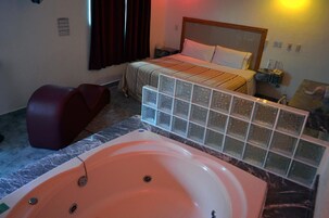 Quarto de hotel com uma banheira grande, uma cama com cabeceira de madeira, uma poltrona vermelha e piso de mármore.