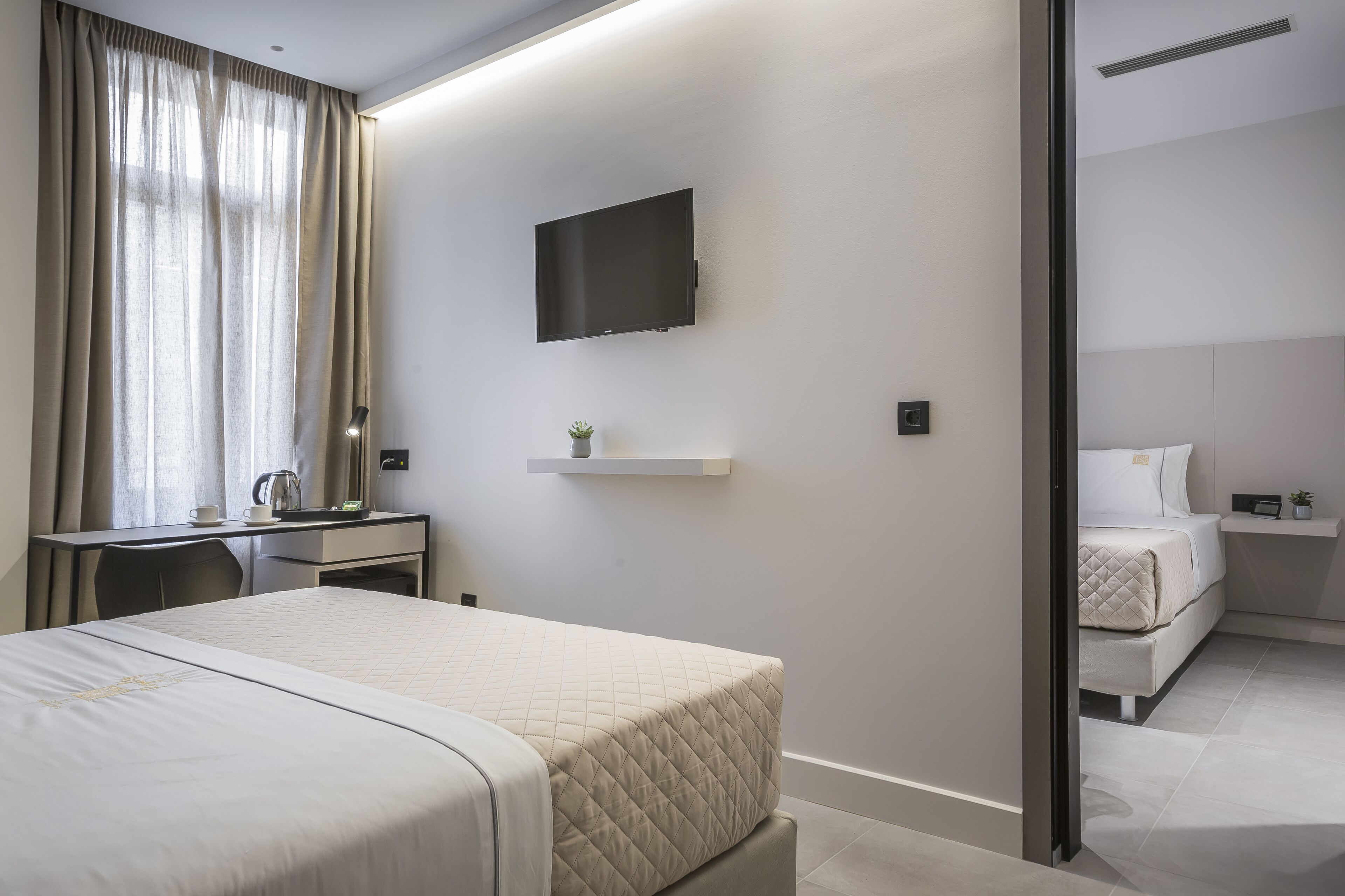 Quarto de hotel com cama, escrivaninha, cadeira, TV fixada na parede e uma pequena estante com uma planta.
